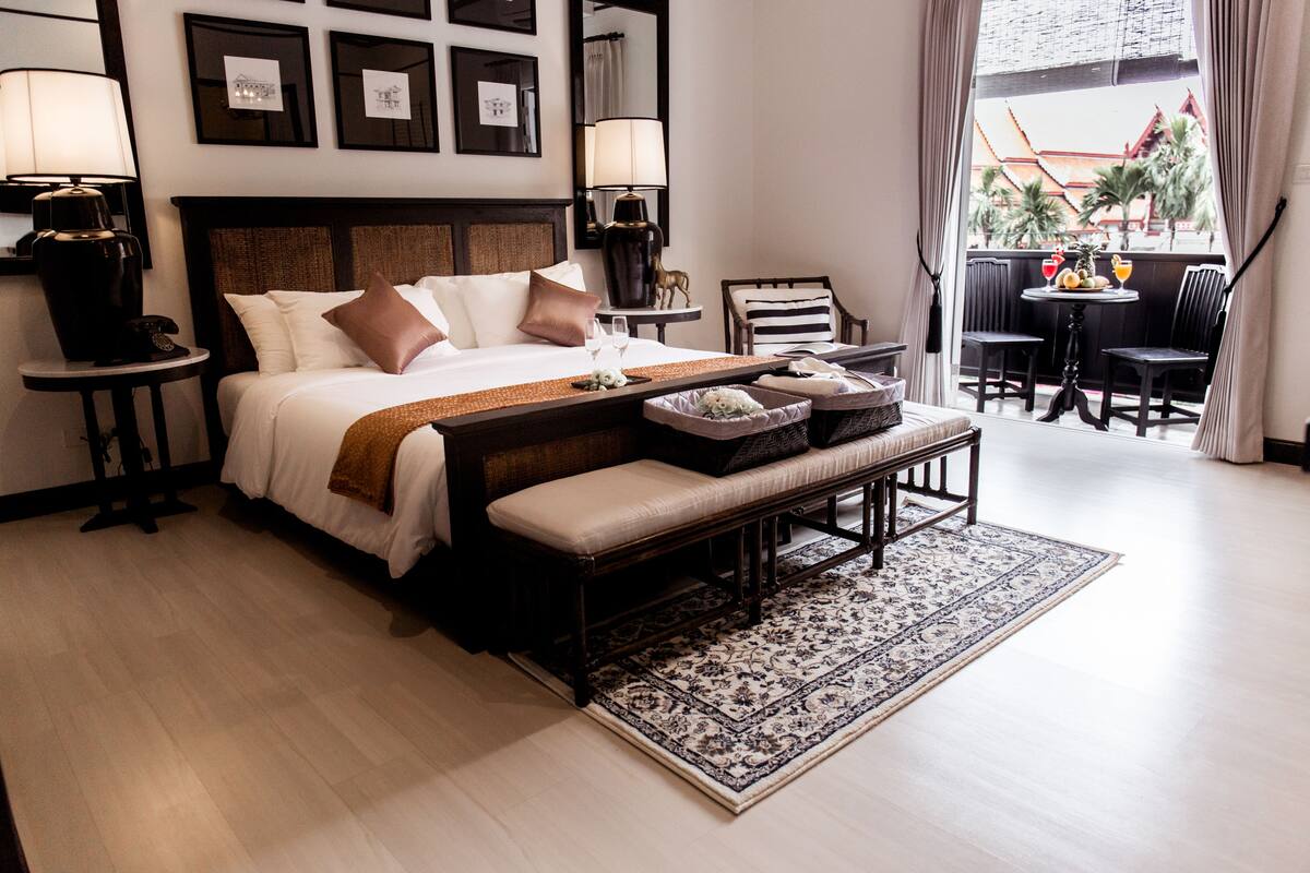 Quarto de hotel com uma cama grande, abajures, um banco, uma mesinha com frutas e bebidas, e vista para o exterior.