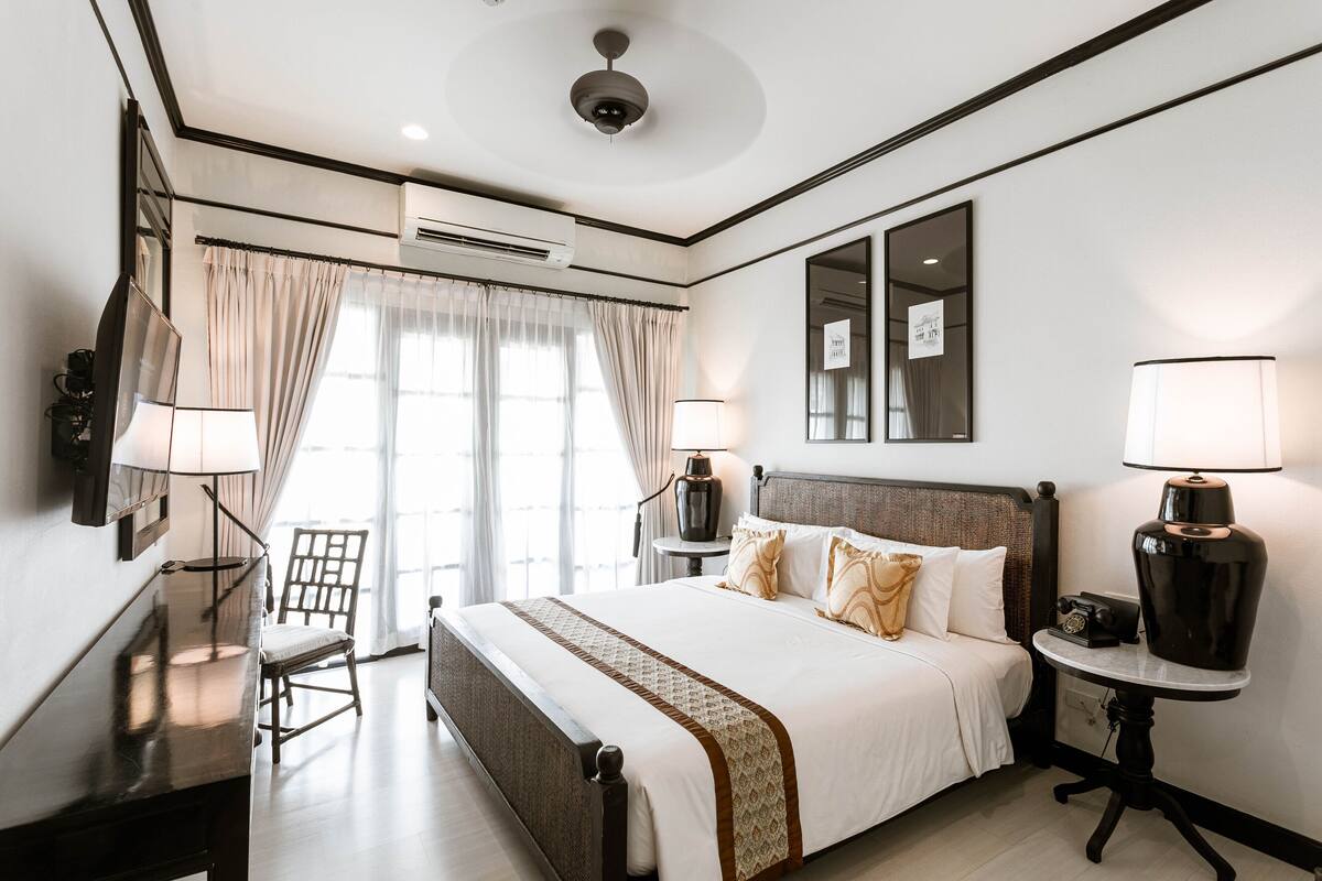 Quarto de hotel com uma cama grande, uma escrivaninha, uma cadeira, uma mesa com abajur e um ventilador de teto.