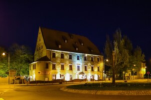 Um edifício histórico com várias janelas e uma entrada imponente, rodeado por árvores e postes de iluminação.