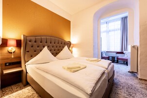 Quarto de hotel com uma cama grande, uma mesa de cabeceira com abajur, e vista para uma área de refeições por uma porta arqueada.