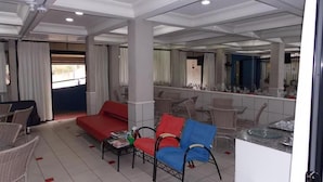 Quarto de hotel com sofá vermelho, poltrona azul e cadeiras de vime. Há uma mesa pequena com revistas e um copo de água. O quarto possui um espelho grande e vista para o exterior através de uma janela com cortinas brancas.