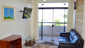 Uma sala de estar com sofá de couro, mesa de centro de vidro, armário de madeira, televisão na parede e porta de vidro deslizante para uma varanda.