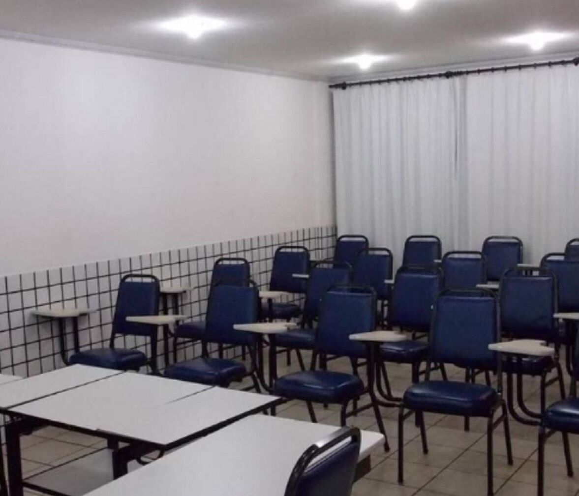 Uma sala de aula com fileiras de cadeiras azuis e mesas brancas, paredes brancas e uma cortina branca.