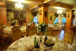 Uma sala de banquetes com mesas redondas preparadas para refeições, com toalhas brancas, cadeiras douradas e arranjos florais.