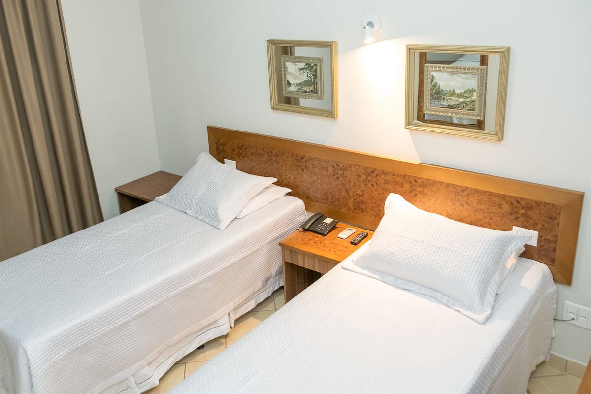 Quarto de hotel com duas camas, cabeceira de madeira, mesa de cabeceira com telefone e controle remoto, e duas fotos emolduradas na parede.