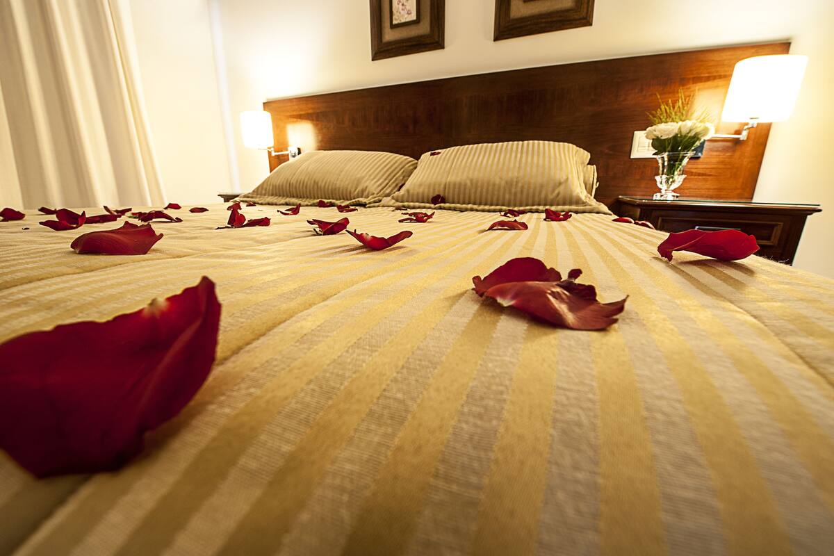 Quarto de hotel com uma cama grande, duas almofadas, uma mesinha de cabeceira com abajur e um vaso com flores.
