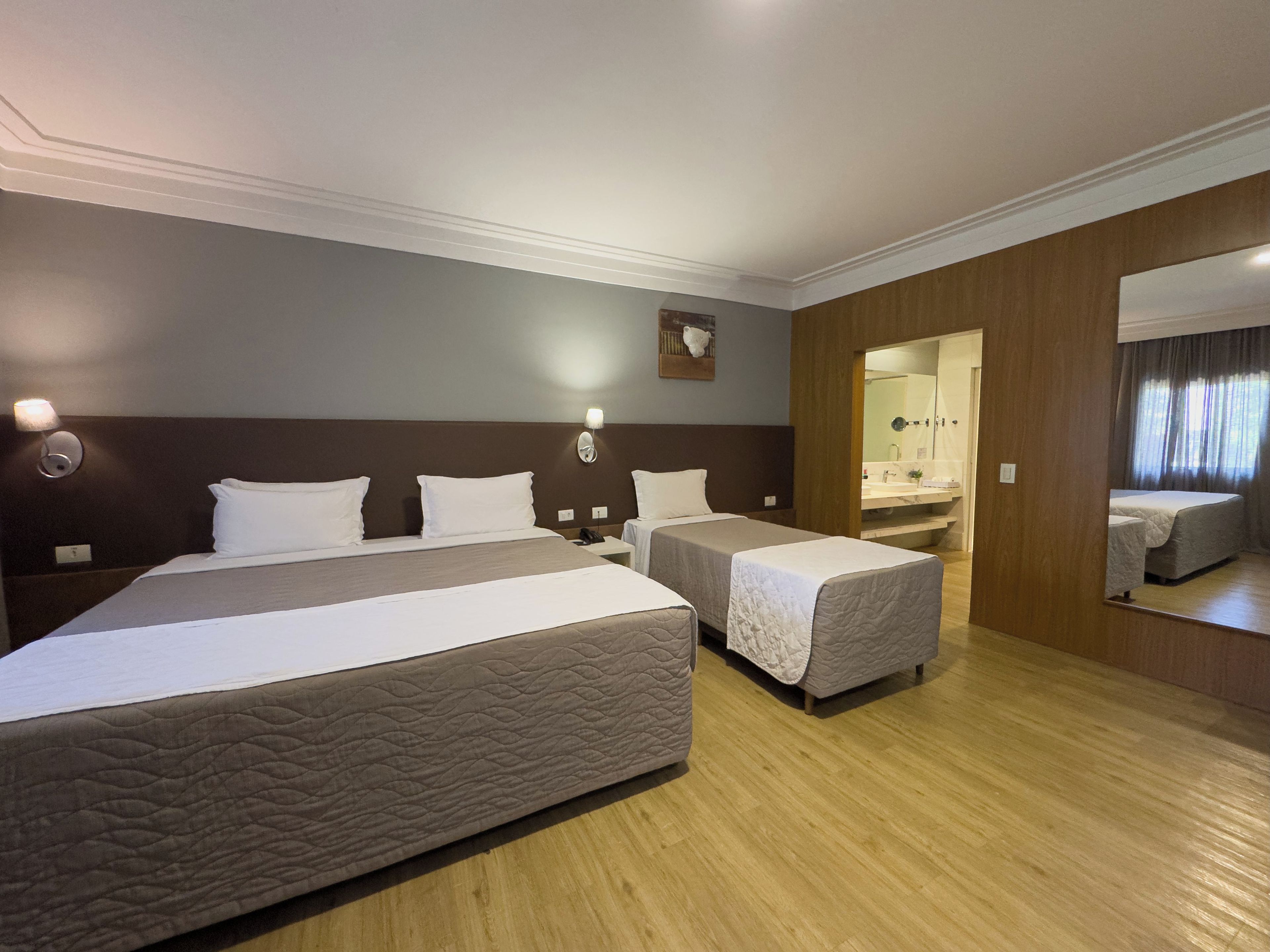 Quarto de hotel com uma cama grande, duas camas menores, piso de madeira, um espelho e um banheiro visível através de uma porta aberta.