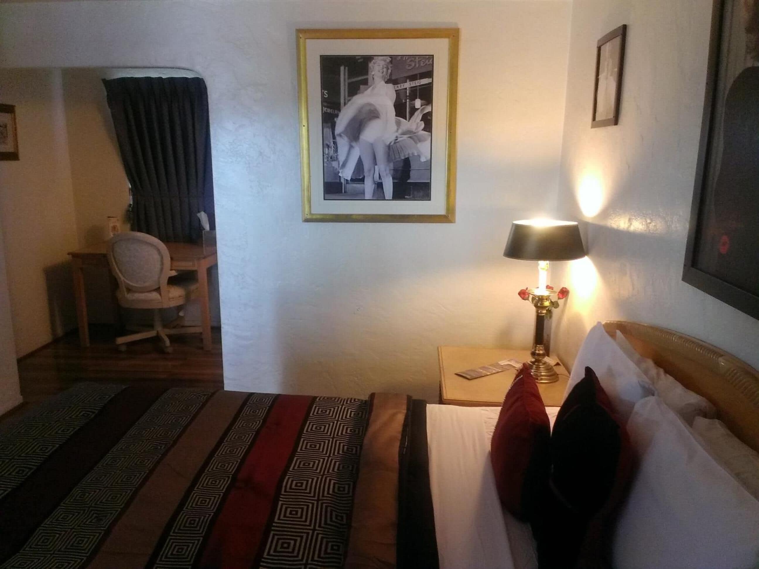 Quarto de hotel com cama, mesa de cabeceira com luminária, uma cadeira, quadro na parede e janela com cortinas.