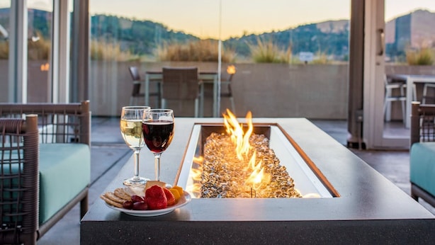 Uma lareira com vista para montanhas, uma mesa com petiscos e duas taças de vinho, e cadeiras de vime.