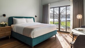 Quarto com cama grande, mesa de cabeceira, luminária e vista para um parque através de uma porta de vidro.