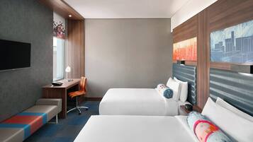 Quarto de hotel com duas camas, uma escrivaninha, uma cadeira, televisão e obras de arte nas paredes.