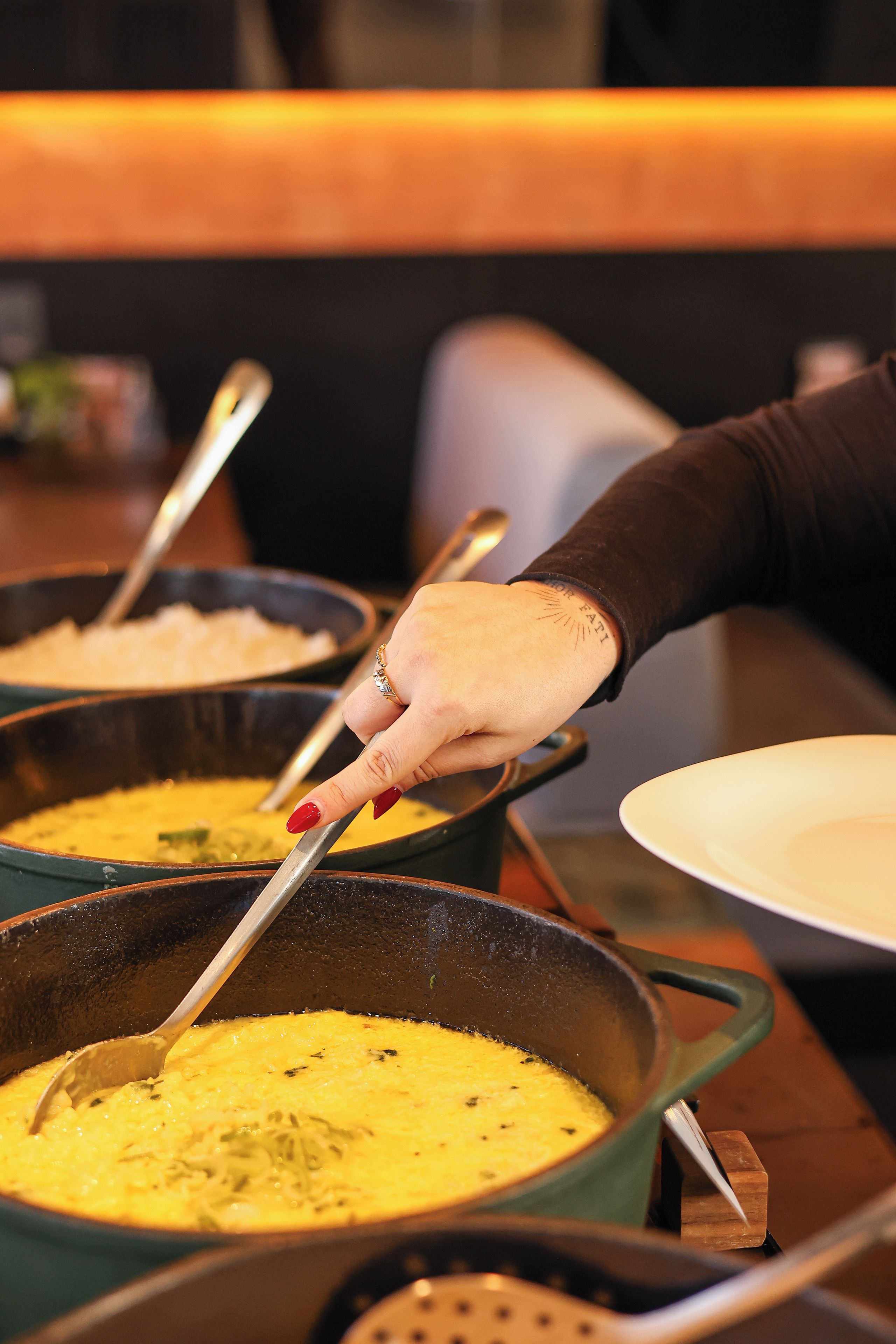 Disposição de buffet com vários recipientes de comida, uma pessoa servindo e uma colher em um dos pratos.
