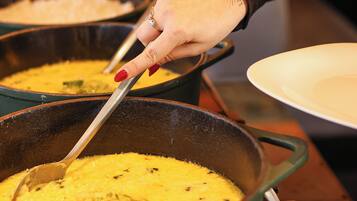 Disposição de buffet com vários recipientes de comida, uma pessoa servindo e uma colher em um dos pratos.