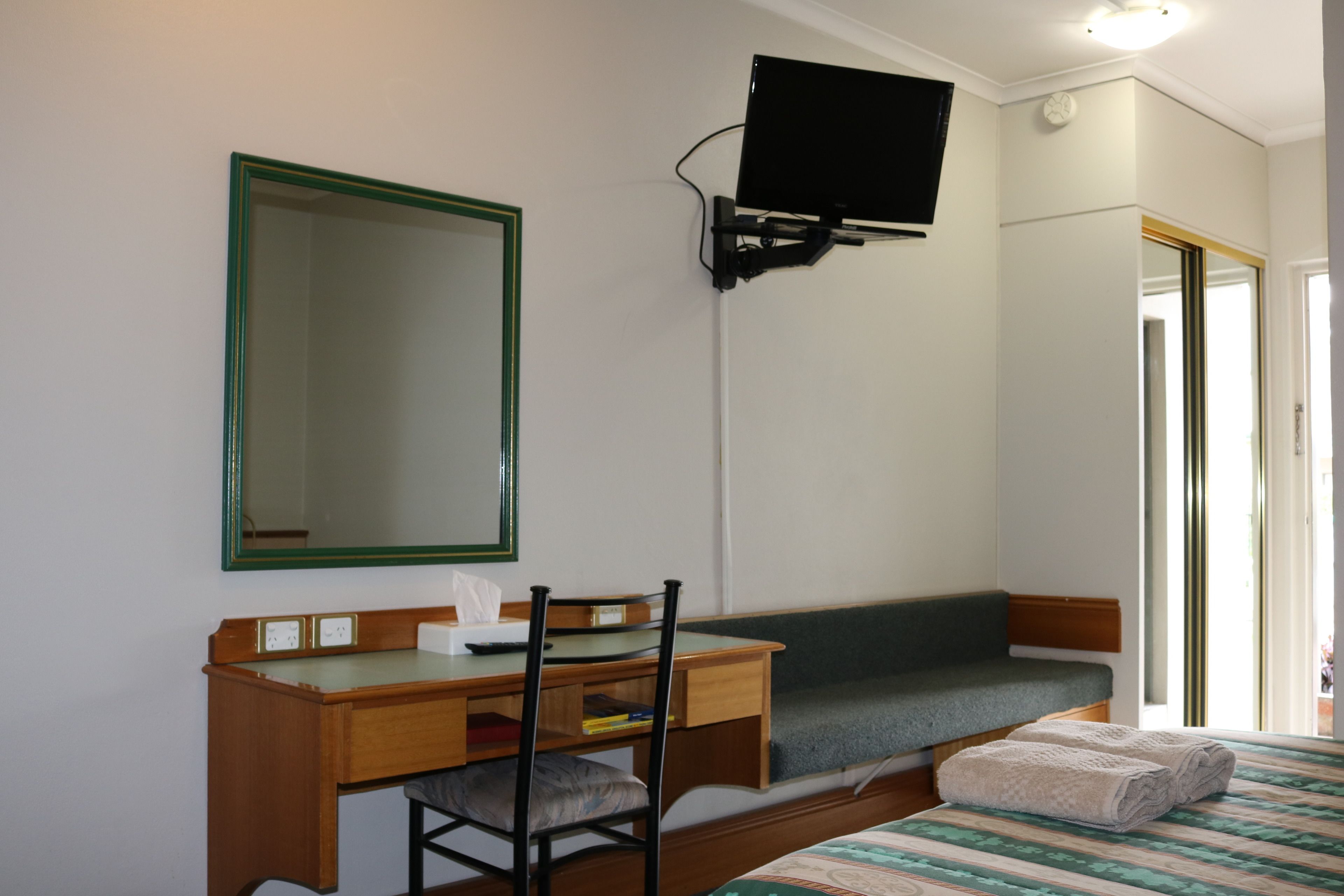 Quarto com cama, escrivaninha com cadeira, televisão na parede e espelho.