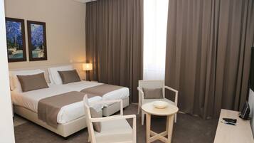 Quarto de hotel com uma cama grande, duas cadeiras, uma mesinha e uma televisão.