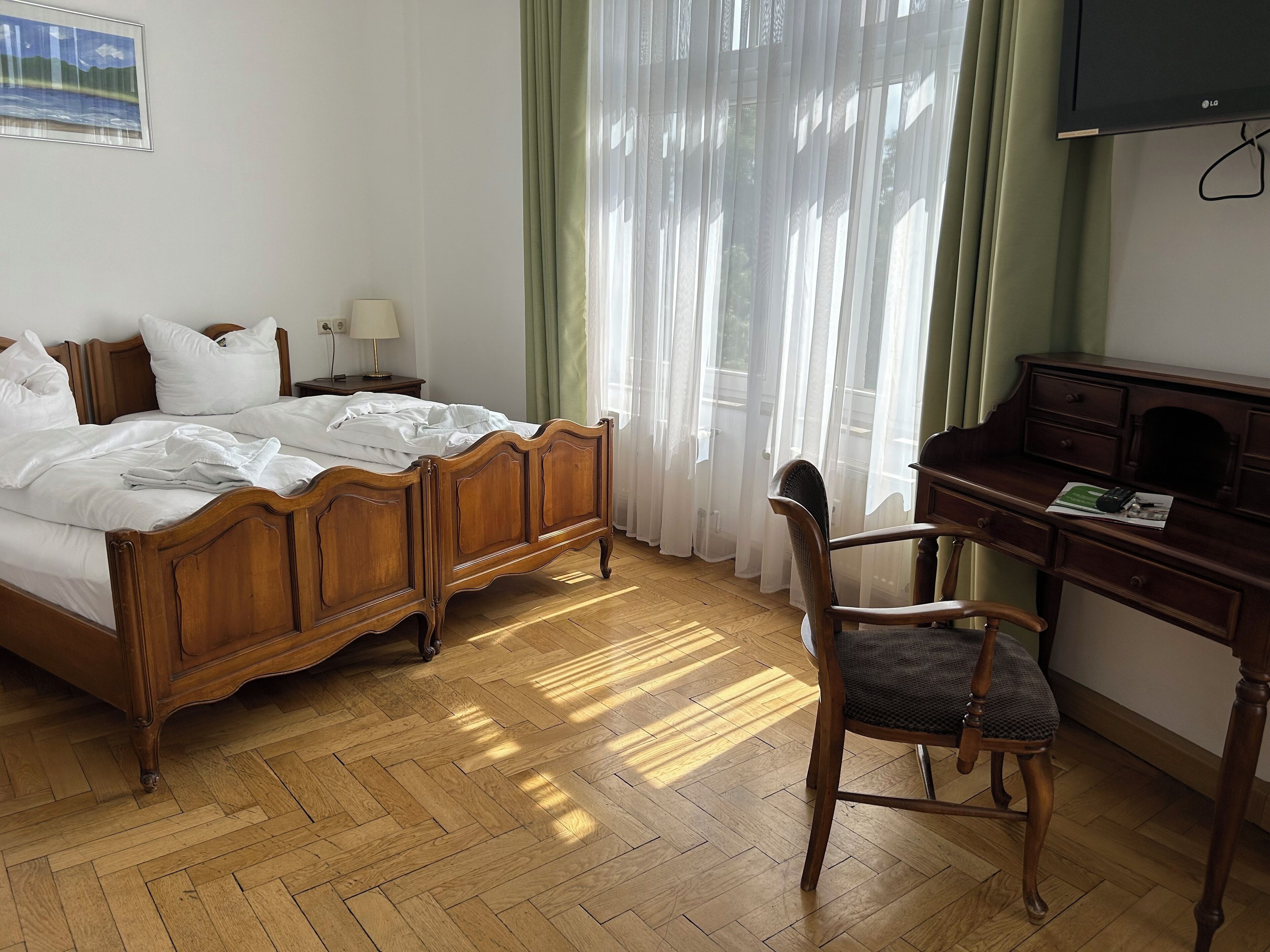 Quarto de hotel com uma cama de madeira, uma escrivaninha com cadeira e uma televisão na parede.
