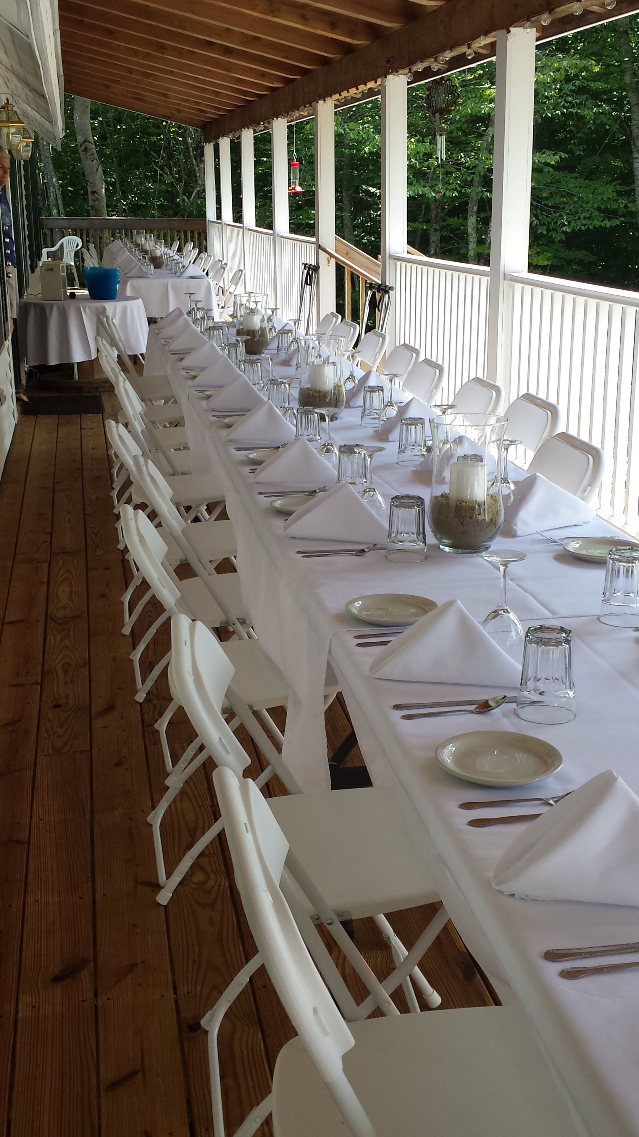 Uma longa mesa posta para um evento formal, com cadeiras brancas e louças, incluindo pratos, copos e talheres.