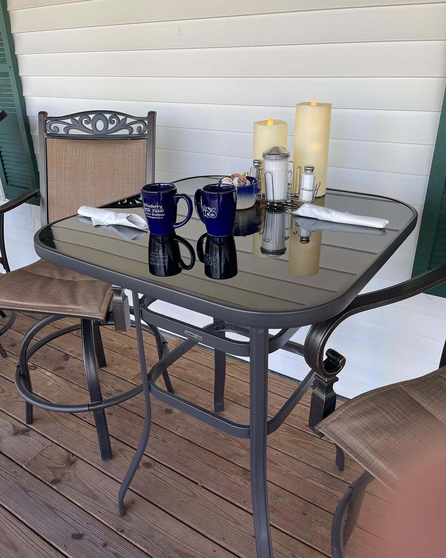Uma mesa com tampo de vidro, duas canecas azuis, um guardanapo branco, uma vela e uma cafeteira sobre um deck de madeira.