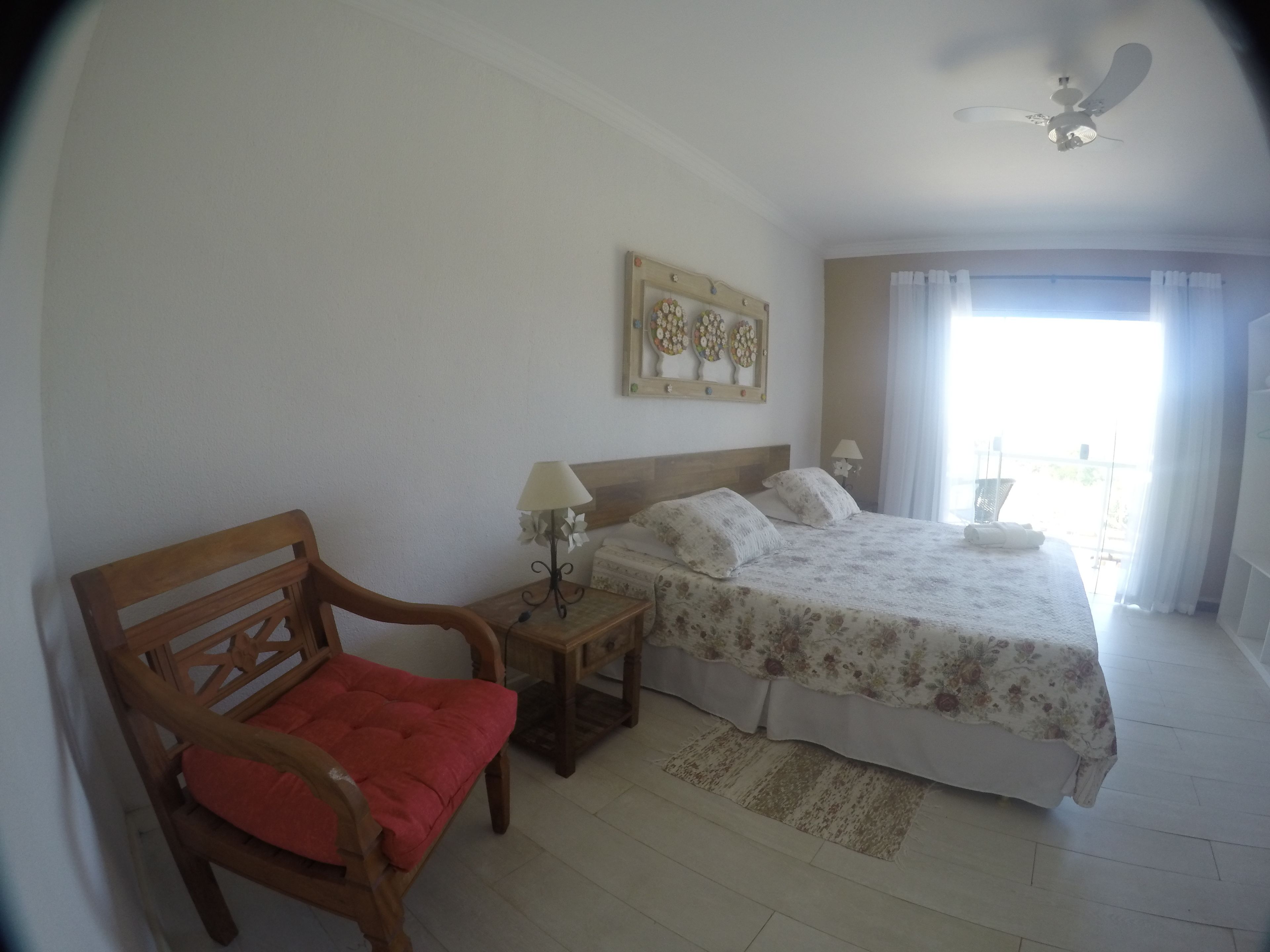 Quarto com cama, uma cadeira, mesa de cabeceira, ventilador de teto e janela com cortinas.