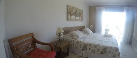 Quarto com cama, uma cadeira, mesa de cabeceira, ventilador de teto e janela com cortinas.