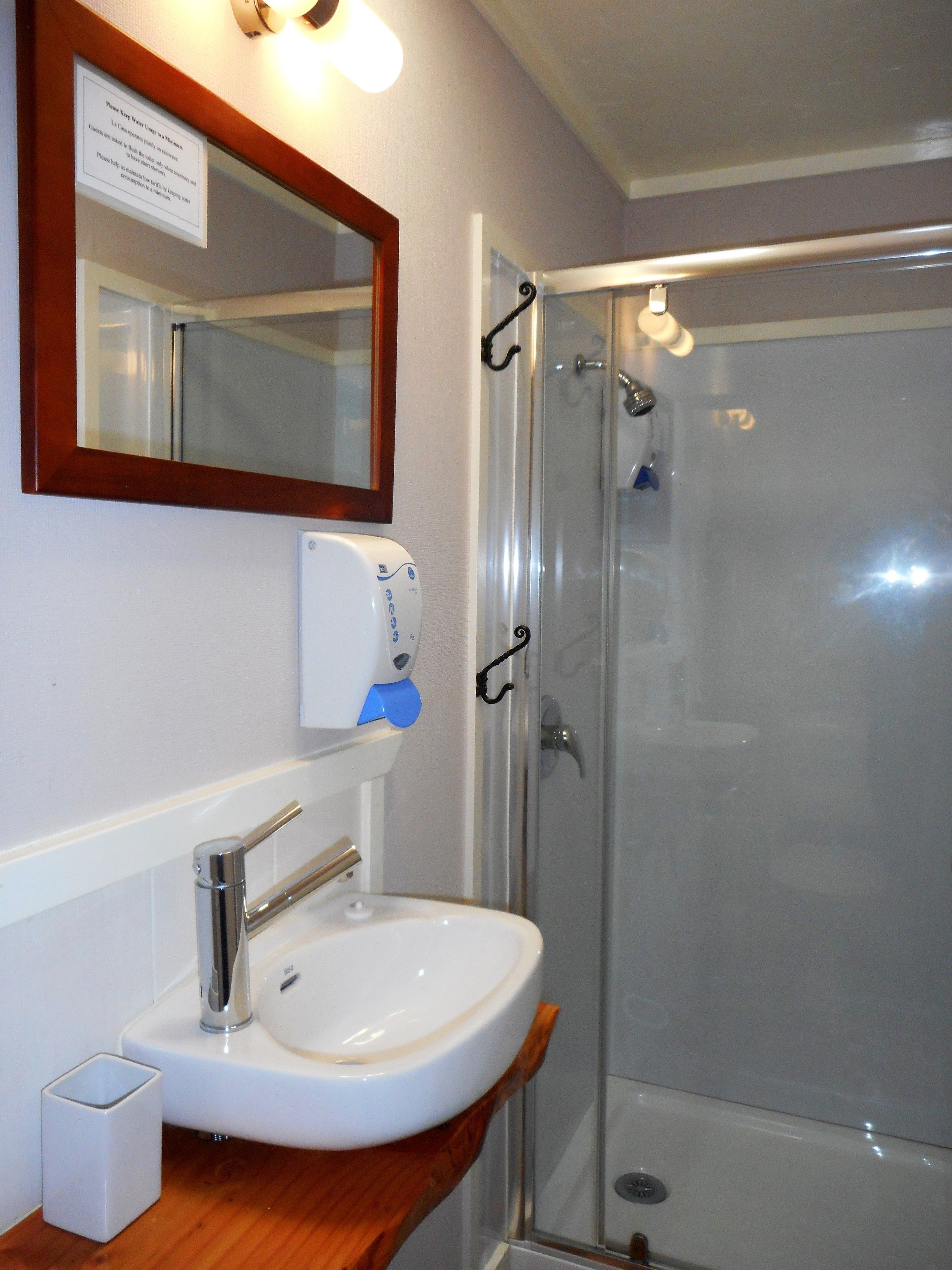 Casa de banho com lavatório branco, espelho, dispensador de sabonete líquido e cabine de duche em vidro.