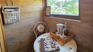 Um quarto aconchegante com paredes de madeira, uma mesa redonda com toalha branca, uma cesta de vime com a etiqueta 'Magazine', uma cadeira de metal, uma janela com vista para árvores e uma prateleira na parede com caixas de madeira.
