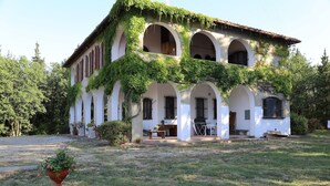 Um edifício branco com janelas em arco e varanda coberta, rodeado por vegetação.