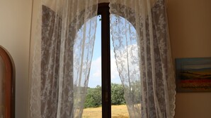 Quarto com uma grande janela coberta por cortinas de renda, um sofá escuro com almofadas decorativas e um quadro na parede.