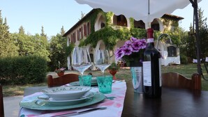 Mesa posta ao ar livre com uma garrafa de vinho, copos e uma tigela sobre uma toalha de mesa.