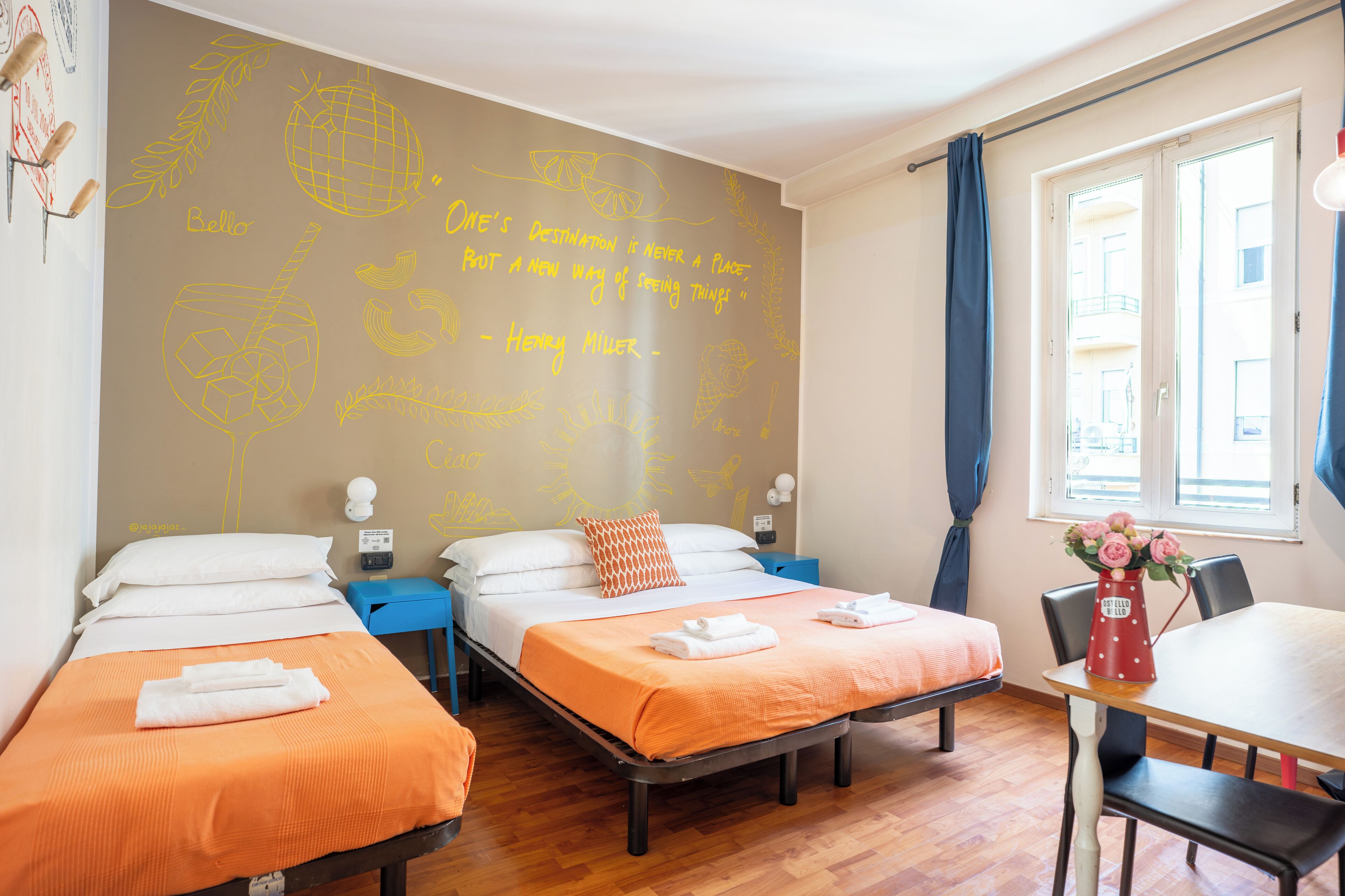Quarto de hotel com duas camas, uma mesa com cadeiras, uma janela com cortinas e uma parede com rabiscos amarelos.