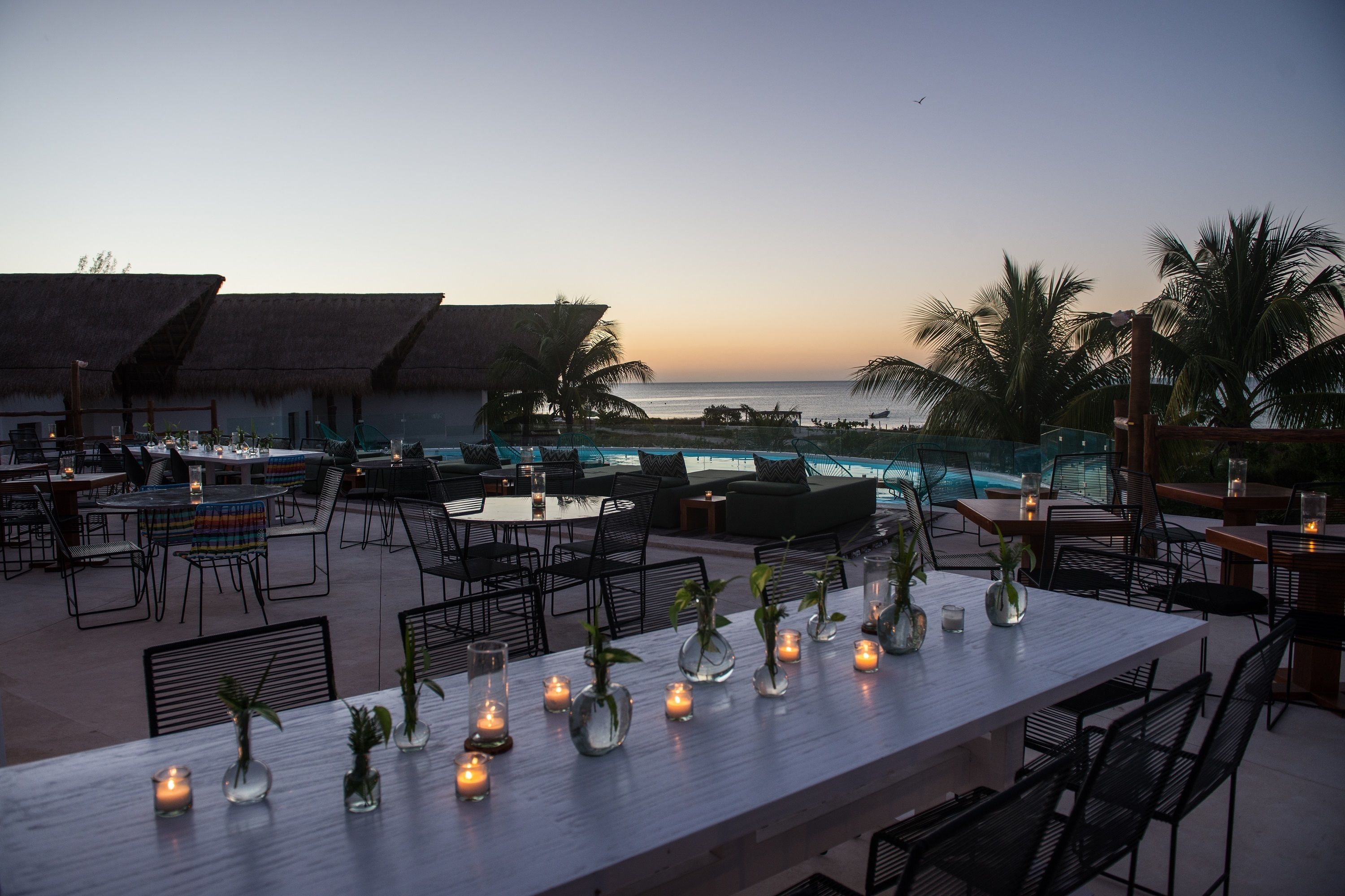 Uma área de refeições ao ar livre com mesas e cadeiras, piscina e vista para o oceano ao pôr do sol.