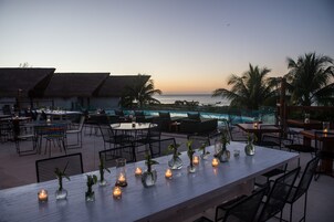 Uma área de refeições ao ar livre com mesas e cadeiras, piscina e vista para o oceano ao pôr do sol.