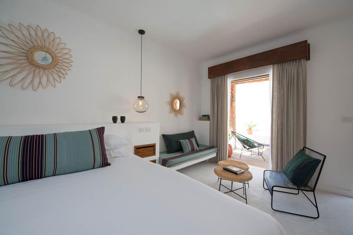 Quarto de hotel com uma cama grande, um sofá, uma cadeira, uma mesa pequena e uma janela com cortinas.