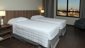 Quarto de hotel com duas camas, uma escrivaninha com luminária, uma cadeira e uma janela grande com vista para a cidade.