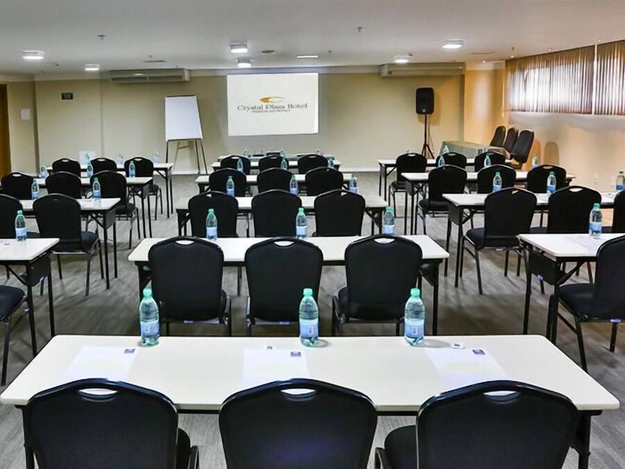 Uma sala de conferências com fileiras de mesas e cadeiras, equipada com garrafas de água, uma tela de projetor e um alto-falante.