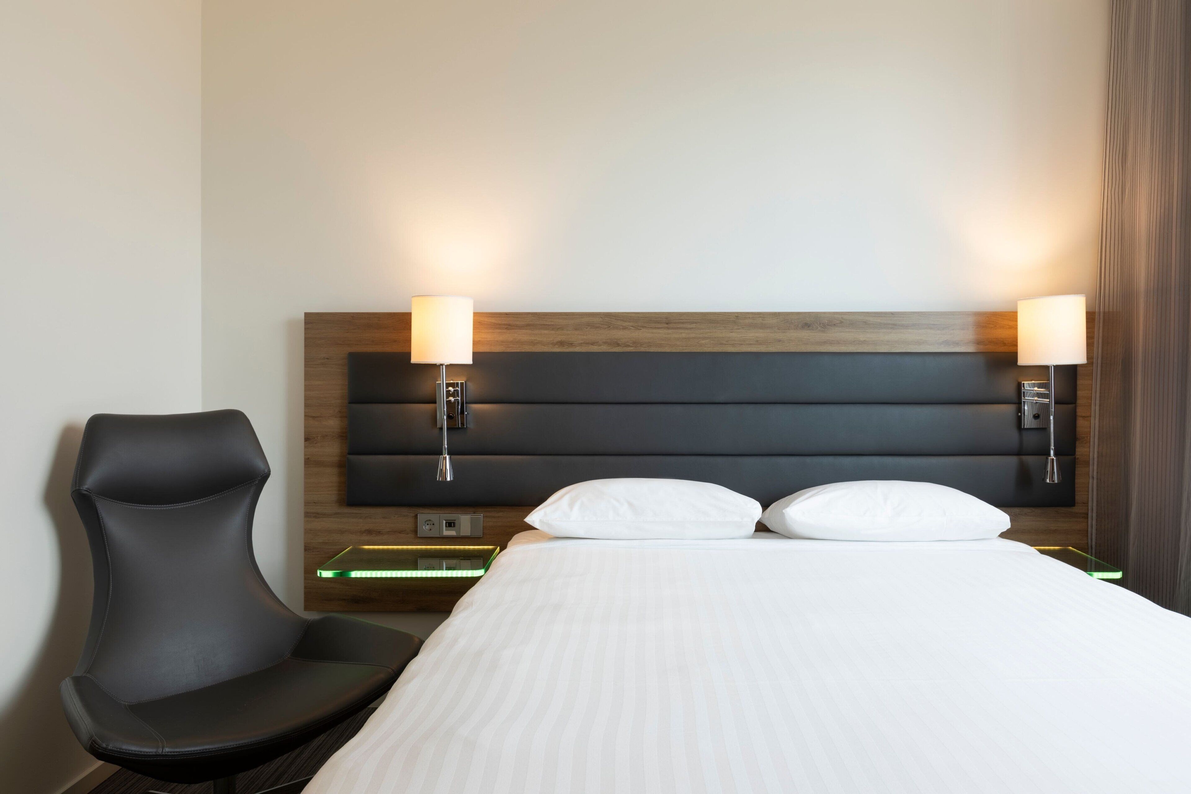 Quarto de hotel com uma cama, duas luminárias de cabeceira, uma poltrona moderna e uma mesa de cabeceira com telefone.