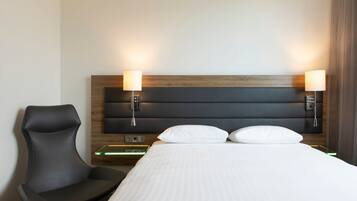 Quarto de hotel com uma cama, duas luminárias de cabeceira, uma poltrona moderna e uma mesa de cabeceira com telefone.