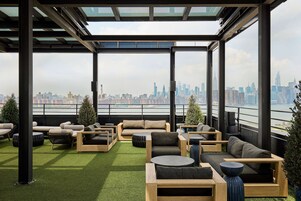 Um lounge no terraço com várias áreas de estar, vista para o horizonte da cidade e um rio ao fundo.