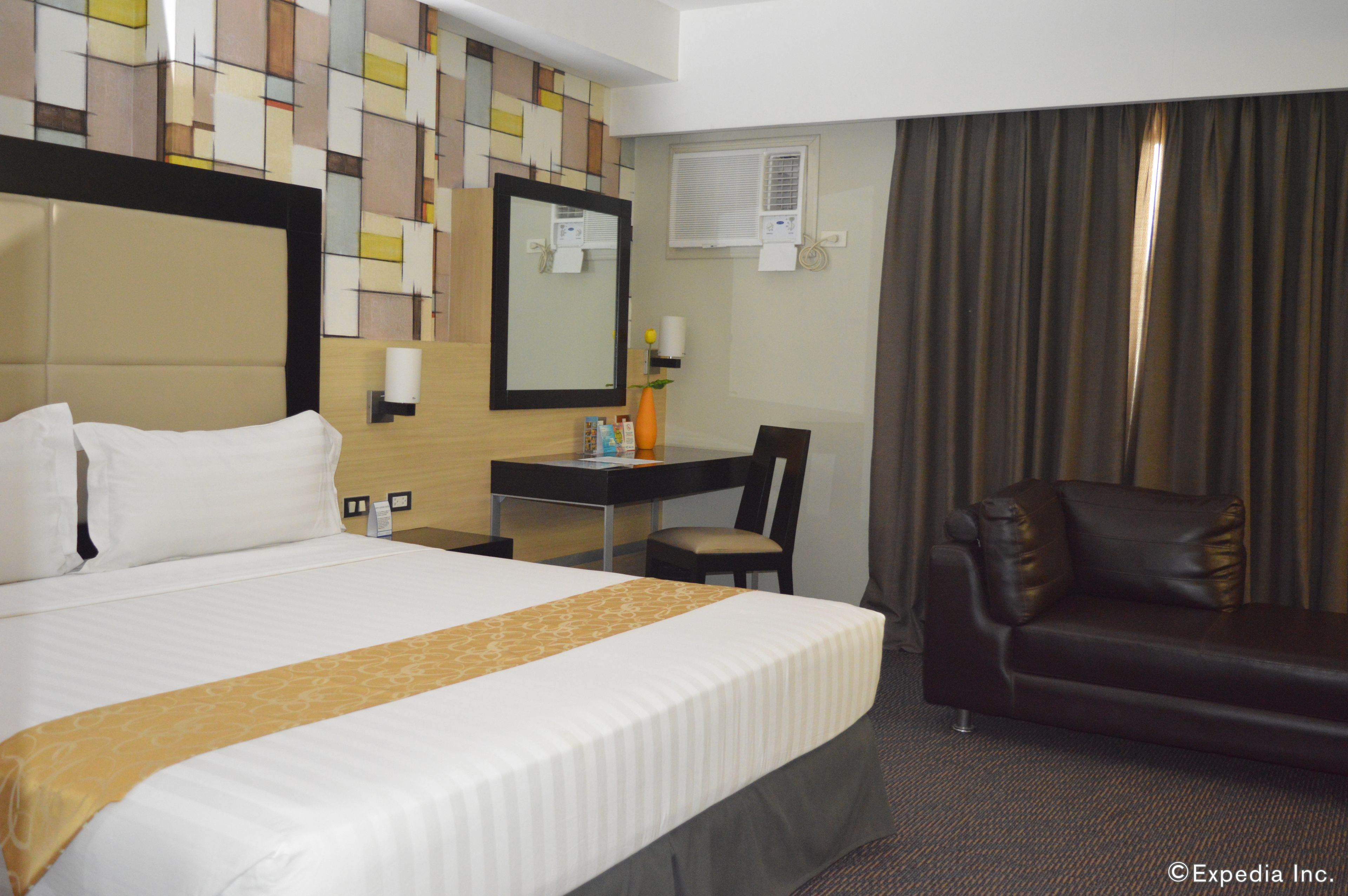 Quarto de hotel com uma cama grande, uma escrivaninha, uma cadeira, um sofá de couro, um espelho e uma janela com cortinas.