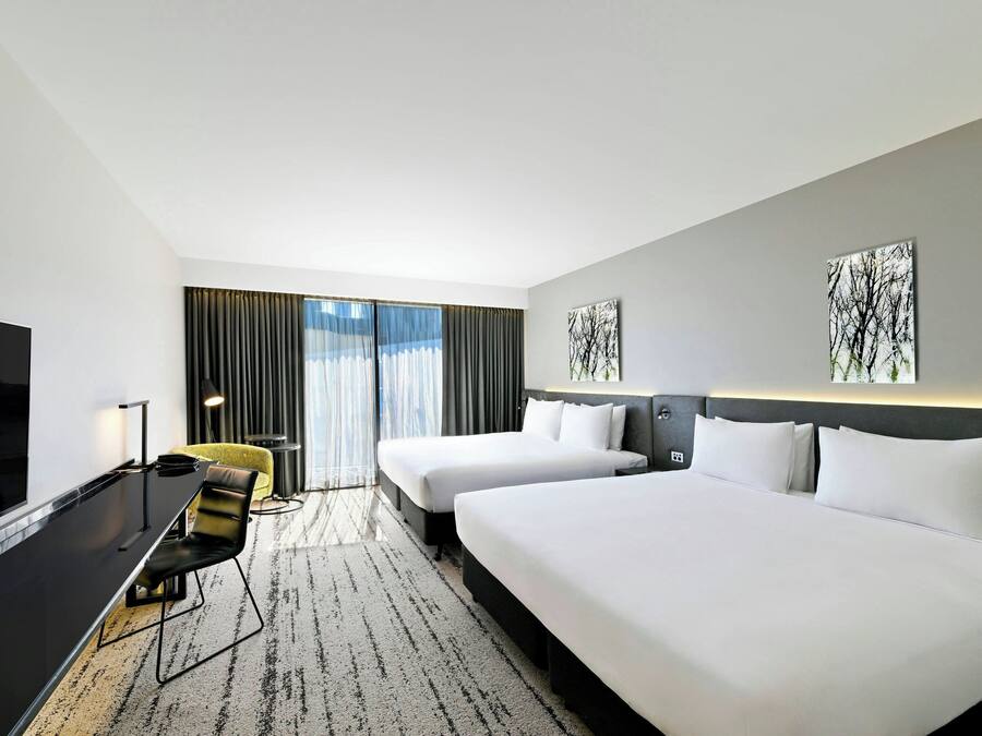 Quarto de hotel com duas camas, uma escrivaninha, televisão e uma janela grande com cortinas.