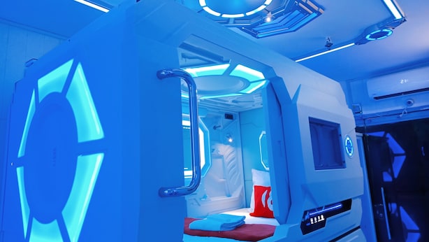 Um quarto futurista com uma luminária circular, uma cama com travesseiro vermelho e branco e um painel de controle na parede.