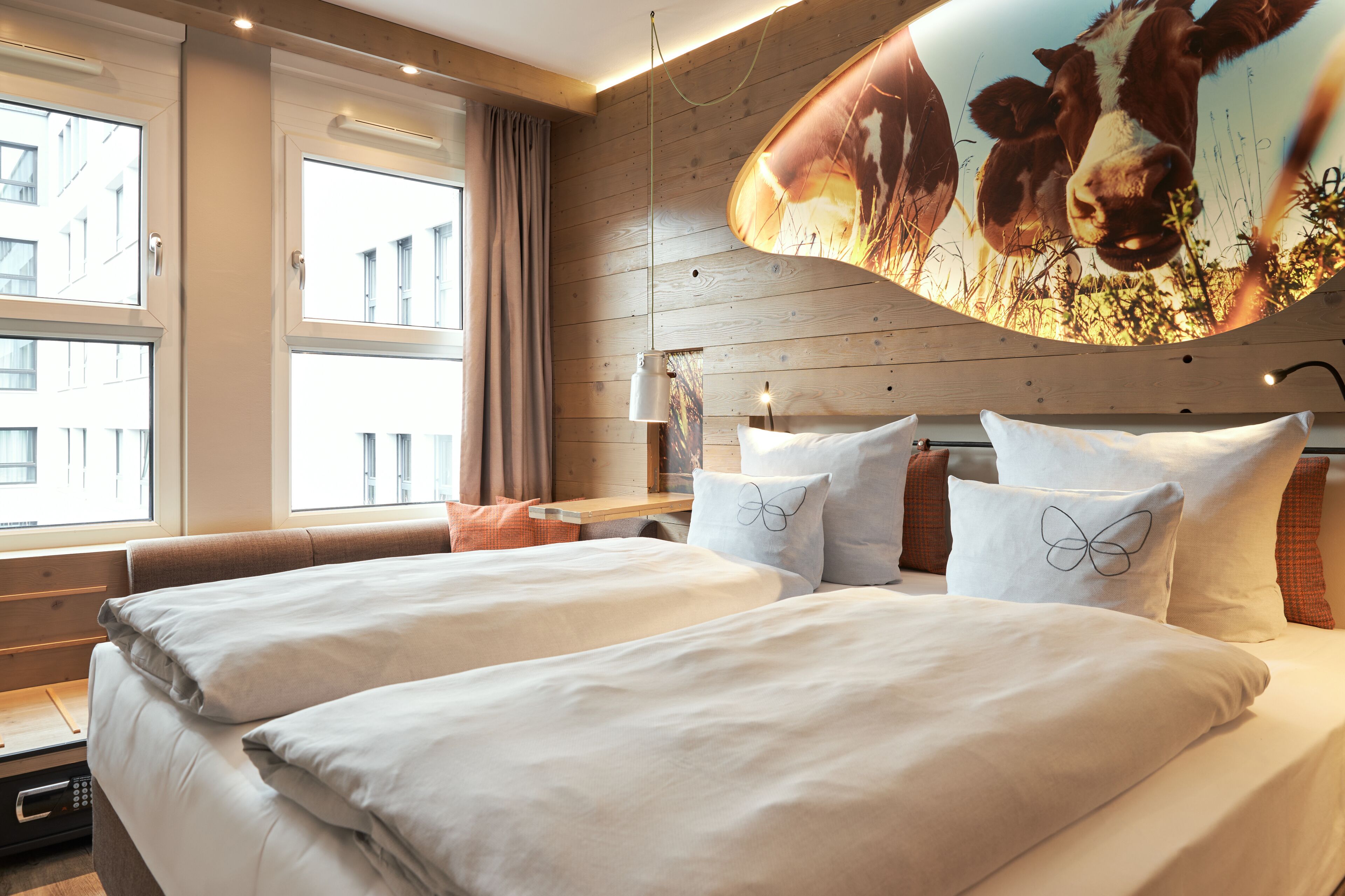Quarto de hotel com duas camas, uma janela grande e um quadro de vaca fixado na parede.