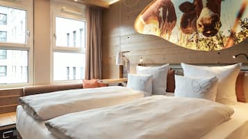 Quarto de hotel com duas camas, uma janela grande e um quadro de vaca fixado na parede.