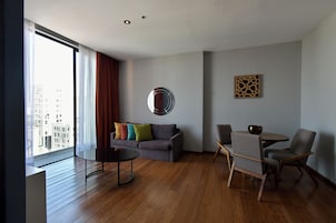 Uma sala de estar moderna com um sofá cinzento, almofadas coloridas, um espelho redondo e um espaço de jantar com duas cadeiras e uma mesa.