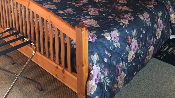 Quarto com cama de madeira, roupa de cama florida, uma cadeira, uma janela com cortinas e um quadro na parede.