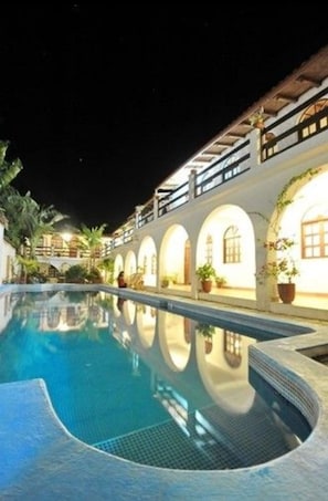 Uma área de piscina à noite, com piscina transparente, janelas em arco e plantas em vasos.