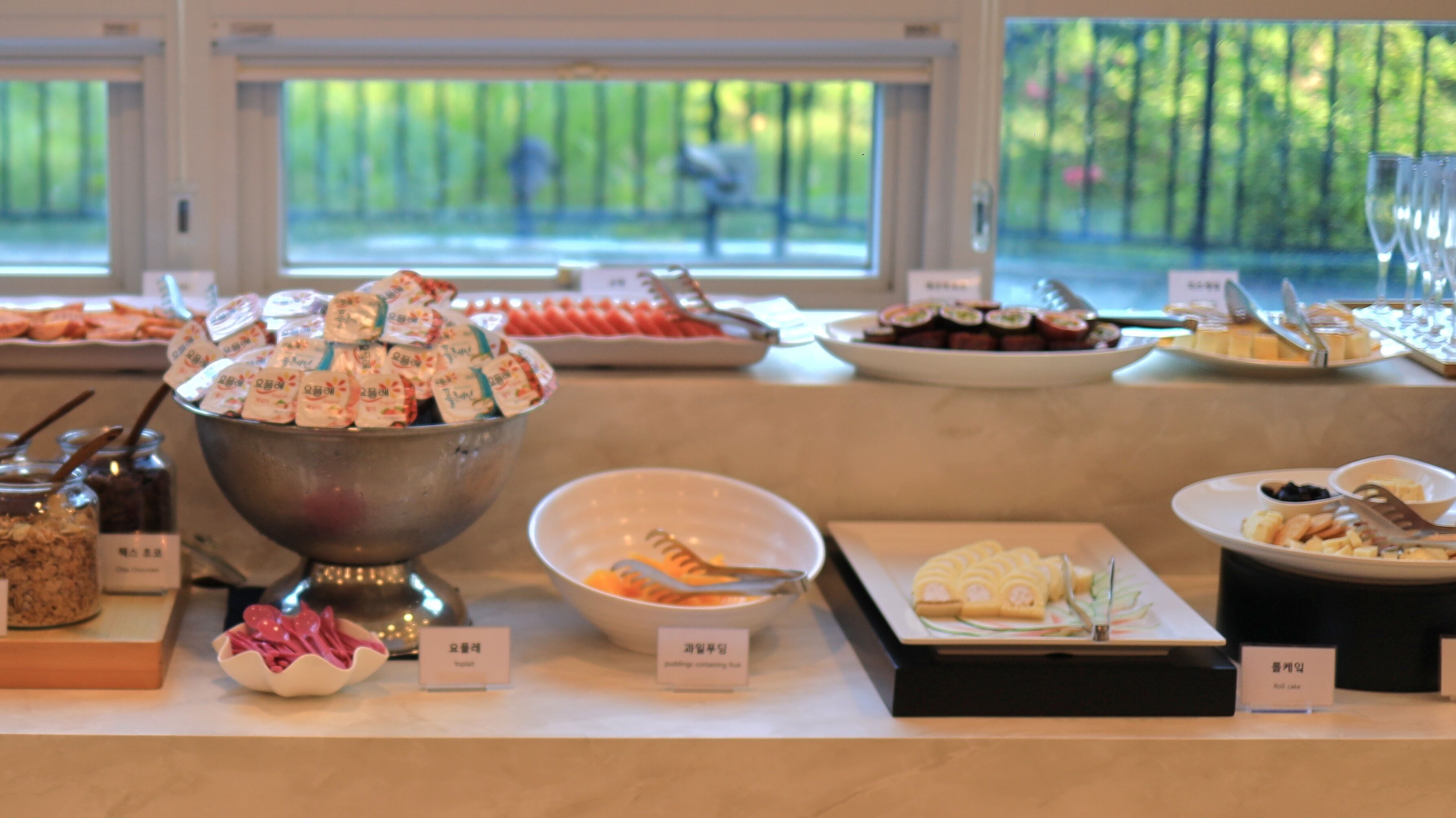 Buffet com diversos alimentos e etiquetas em ambiente bem iluminado com grandes janelas.