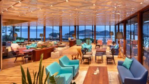 Uma sala de estar ampla com teto de madeira, grandes janelas com vista para o mar e colinas, e diversas opções de assentos, incluindo sofás e poltronas.