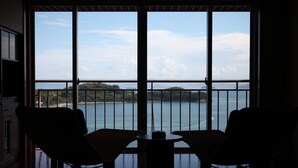 Quarto com uma grande janela com vista para um corpo de água e colinas ao longe. Há duas cadeiras e uma pequena mesa no quarto.