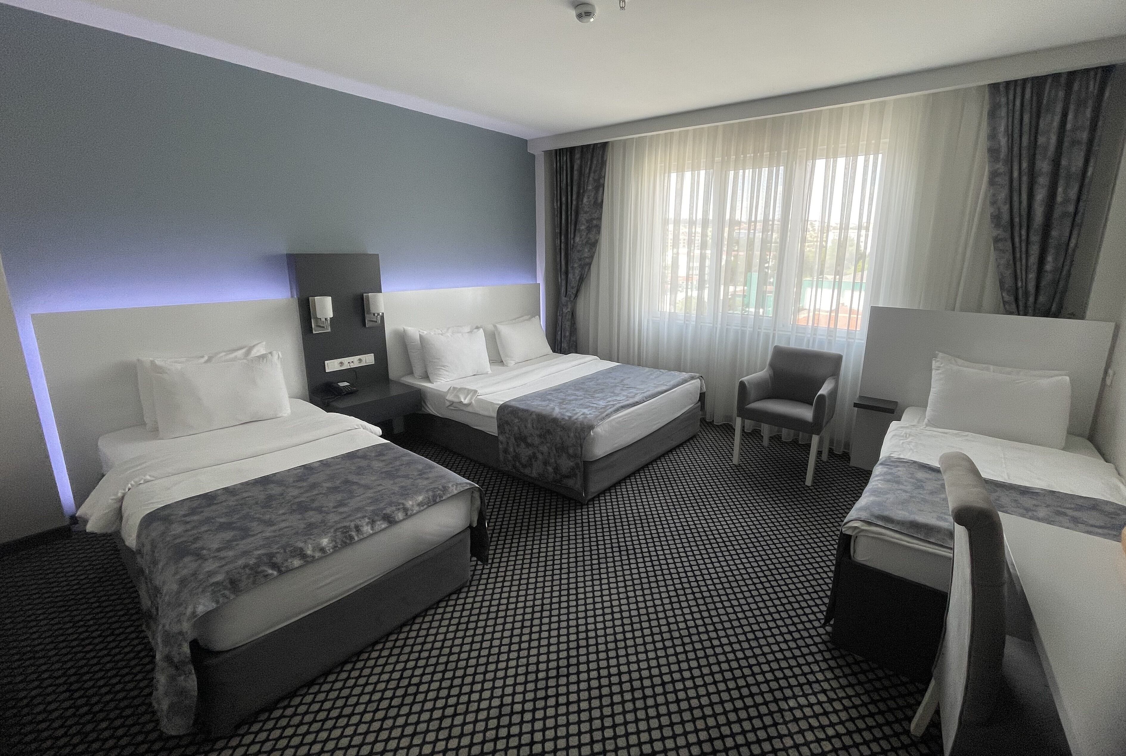 Quarto de hotel com duas camas, uma escrivaninha, uma cadeira e uma janela grande com cortinas.